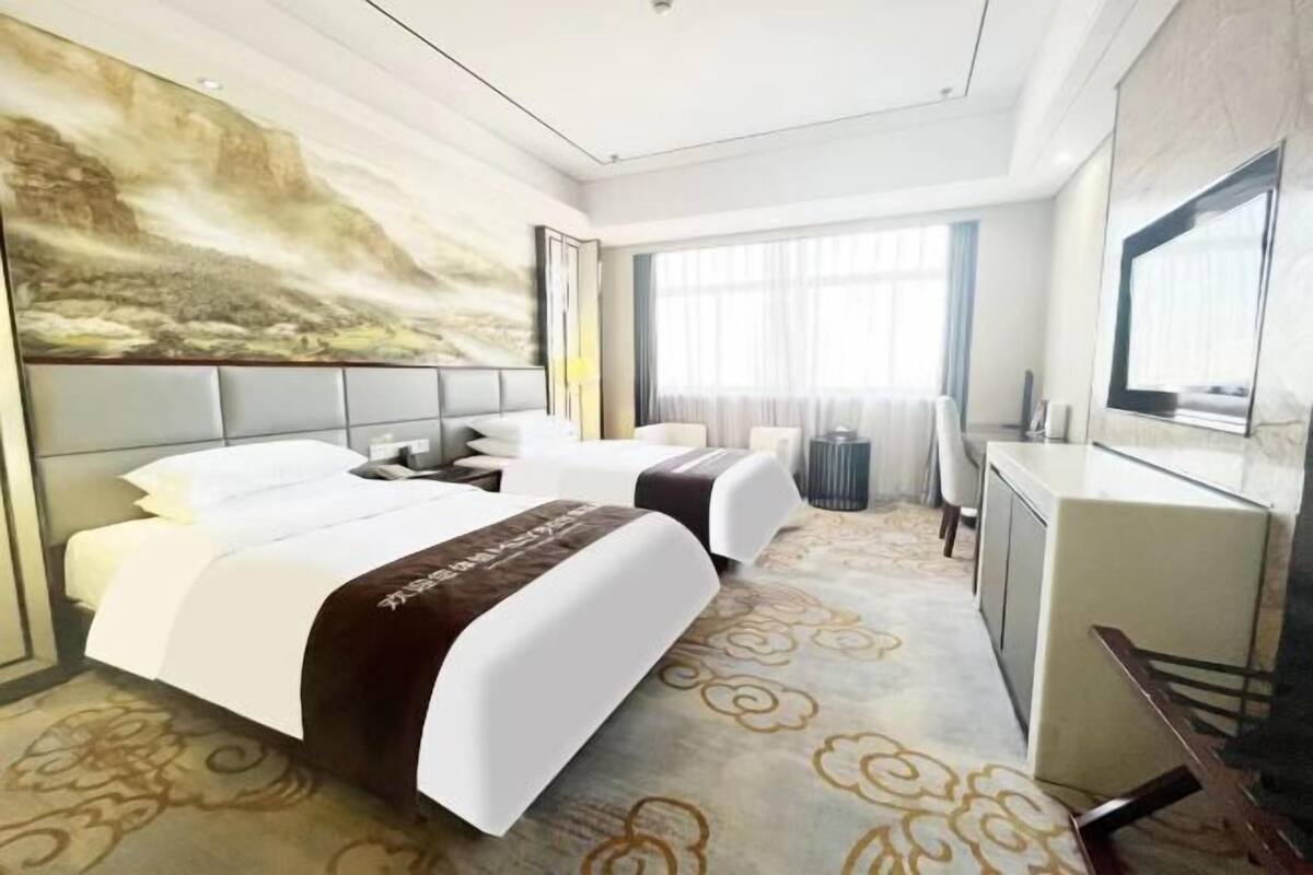Quarto de hotel com duas camas, uma escrivaninha, uma cadeira, uma televisão e um quadro grande na parede.