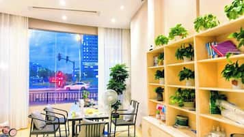 Quarto com uma mesa de jantar, cadeiras, um ventilador e vista para uma rua da cidade. Há prateleiras com plantas e livros.