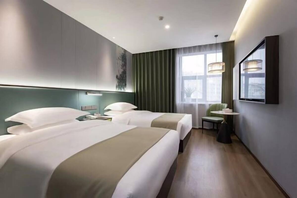 Quarto de hotel com duas camas, uma mesinha, uma cadeira, uma janela com cortinas e uma TV fixada na parede.