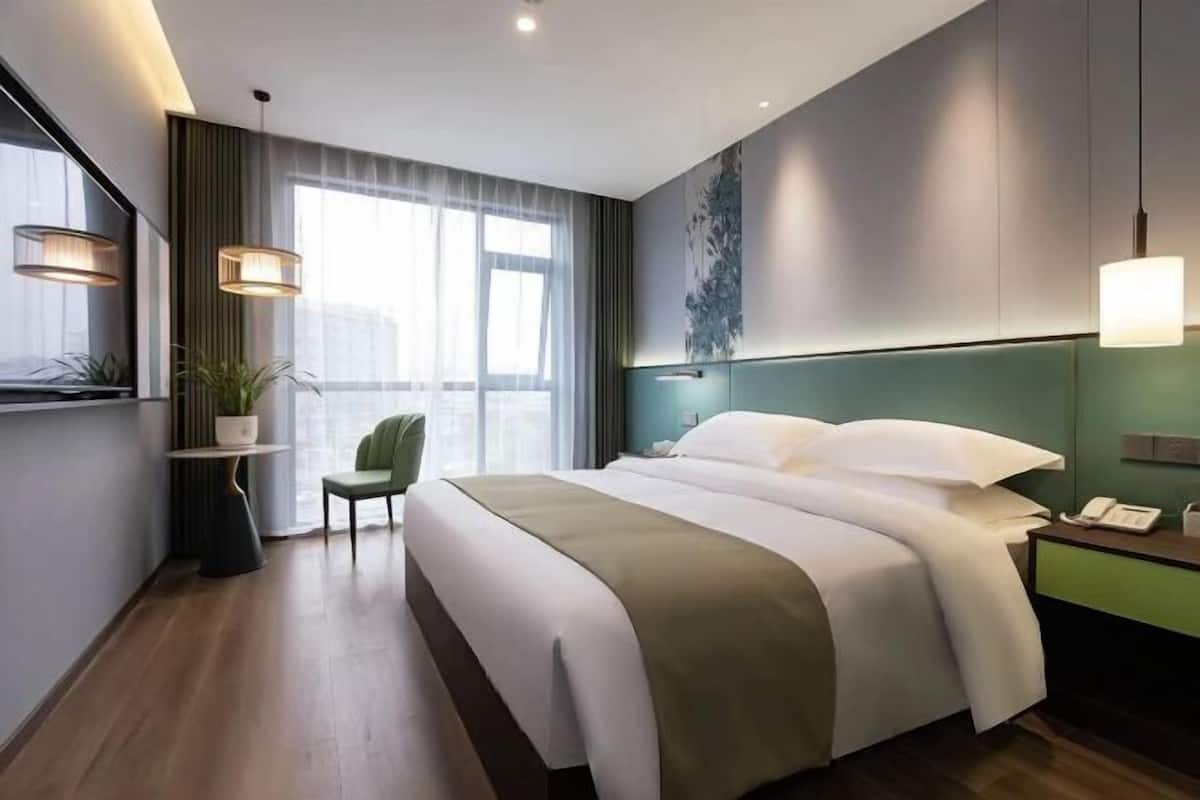 Quarto de hotel com uma cama grande, uma mesa de cabeceira com telefone, uma cadeira, uma janela com cortinas e uma obra de arte na parede.