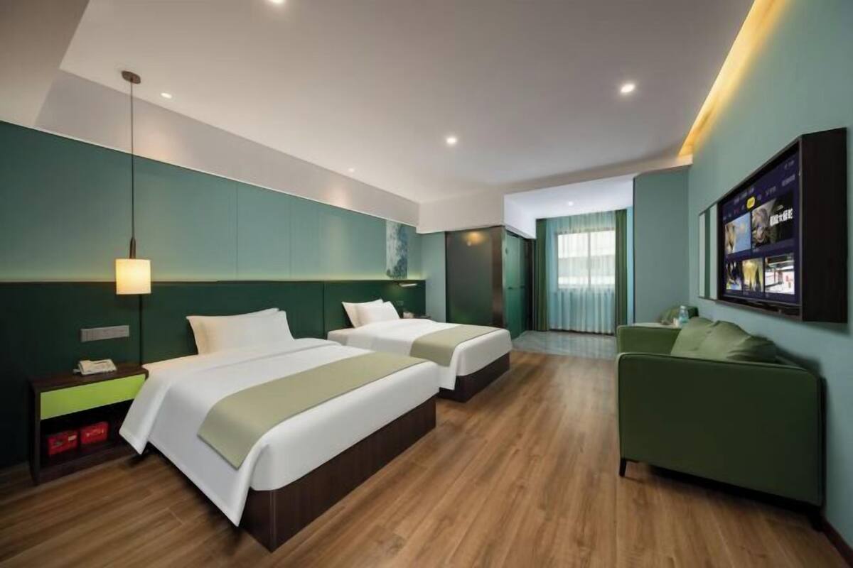 Quarto de hotel com duas camas, um sofá verde, uma televisão e piso de madeira.