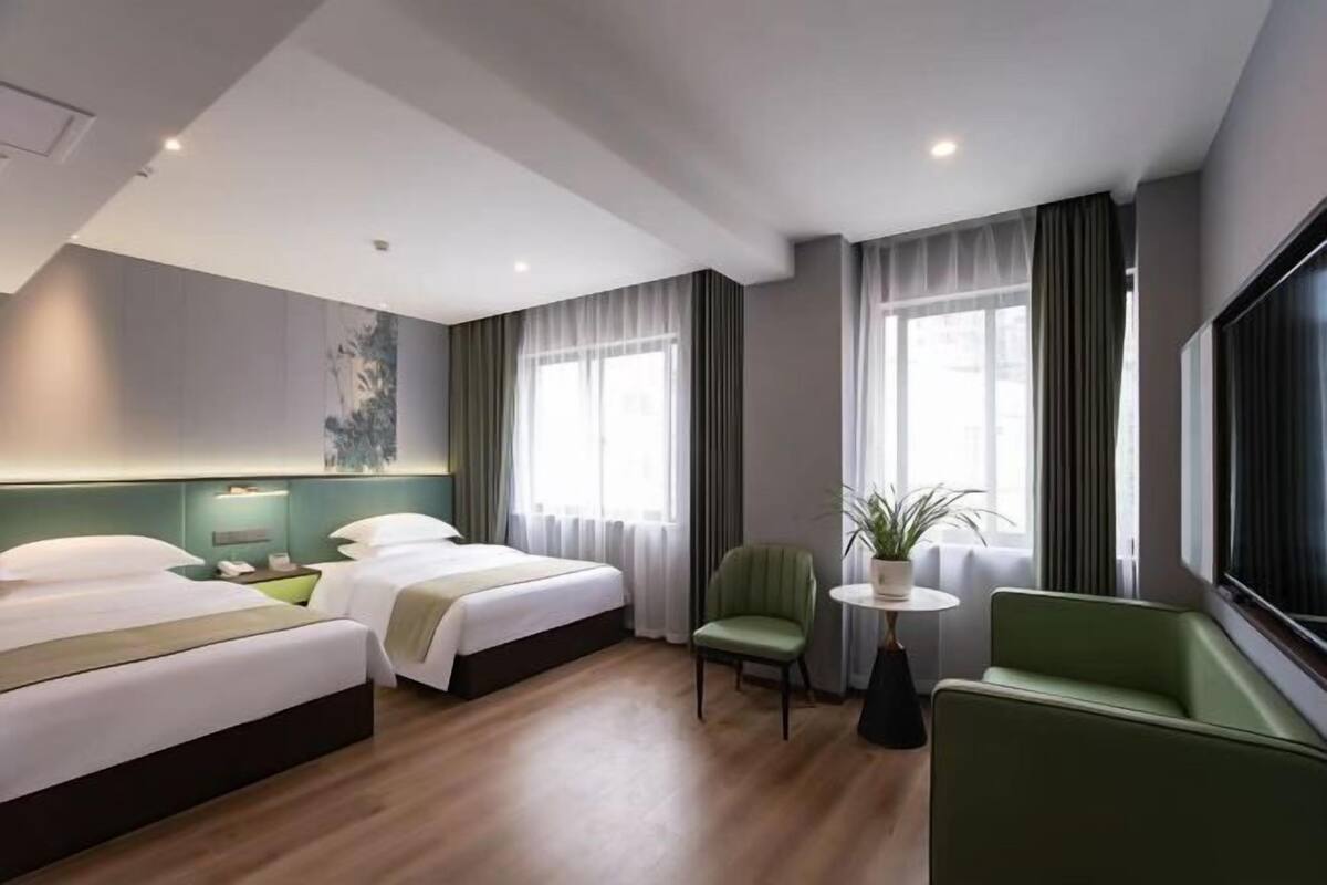 Quarto de hotel com duas camas, uma poltrona verde, uma mesa pequena com uma planta e grandes janelas com cortinas.