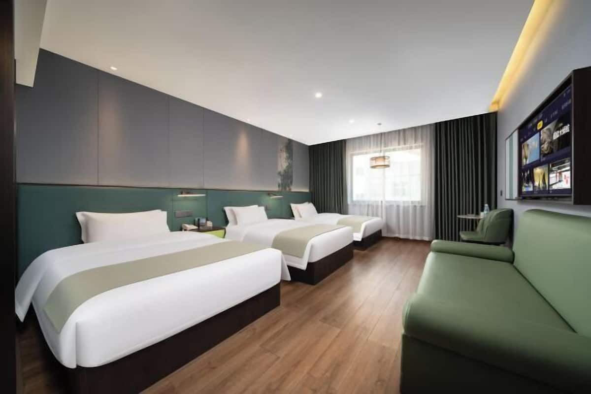 Quarto de hotel com três camas, um sofá verde, uma mesinha e uma estante.