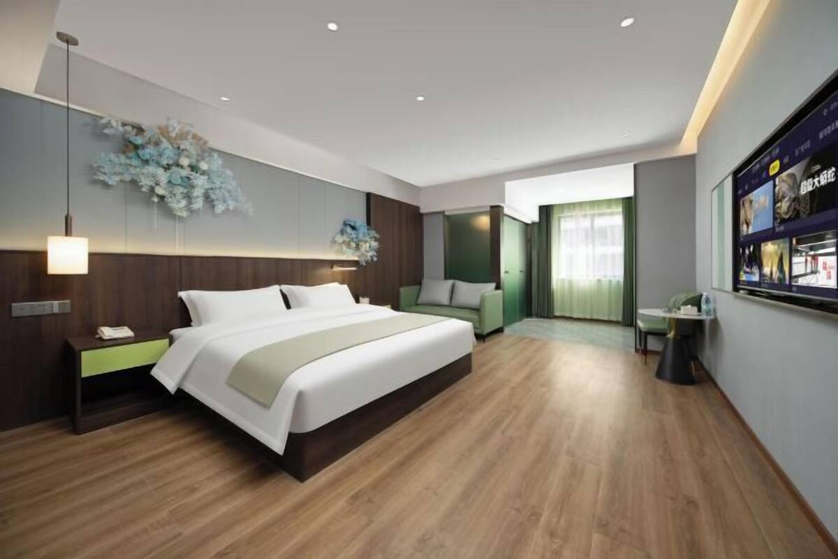Quarto de hotel moderno com uma cama grande, uma mesinha de cabeceira, um sofá verde, uma pequena mesa e uma televisão fixada na parede.