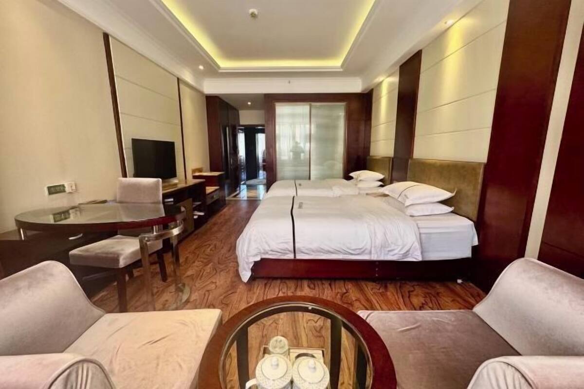 Quarto de hotel com uma cama, duas mesas de cabeceira, uma televisão, um espaço de estar com uma mesa e cadeiras, e um espelho grande.