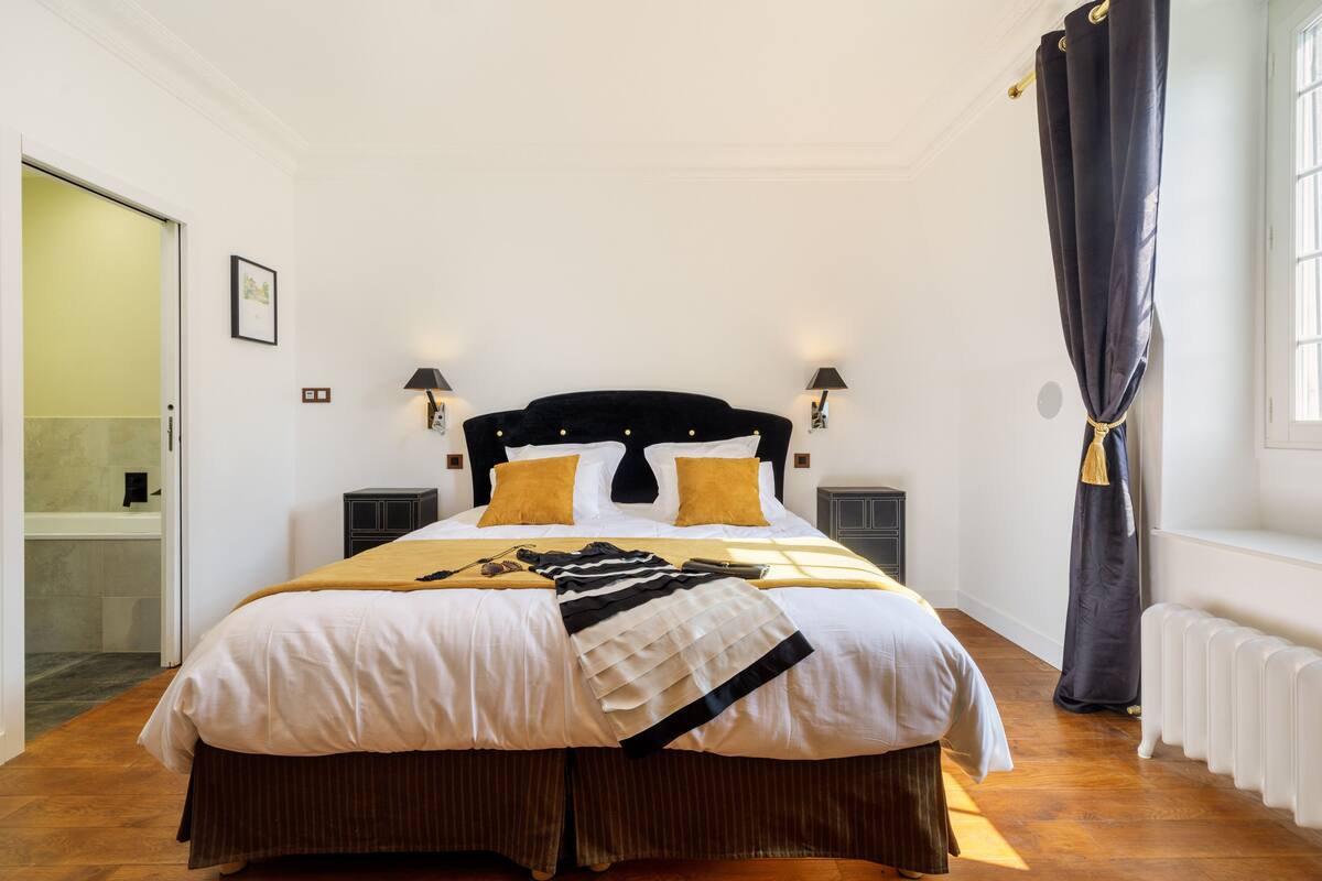 Cama bem arrumada com manta listrada, duas luminárias de cabeceira e uma janela grande com cortinas.