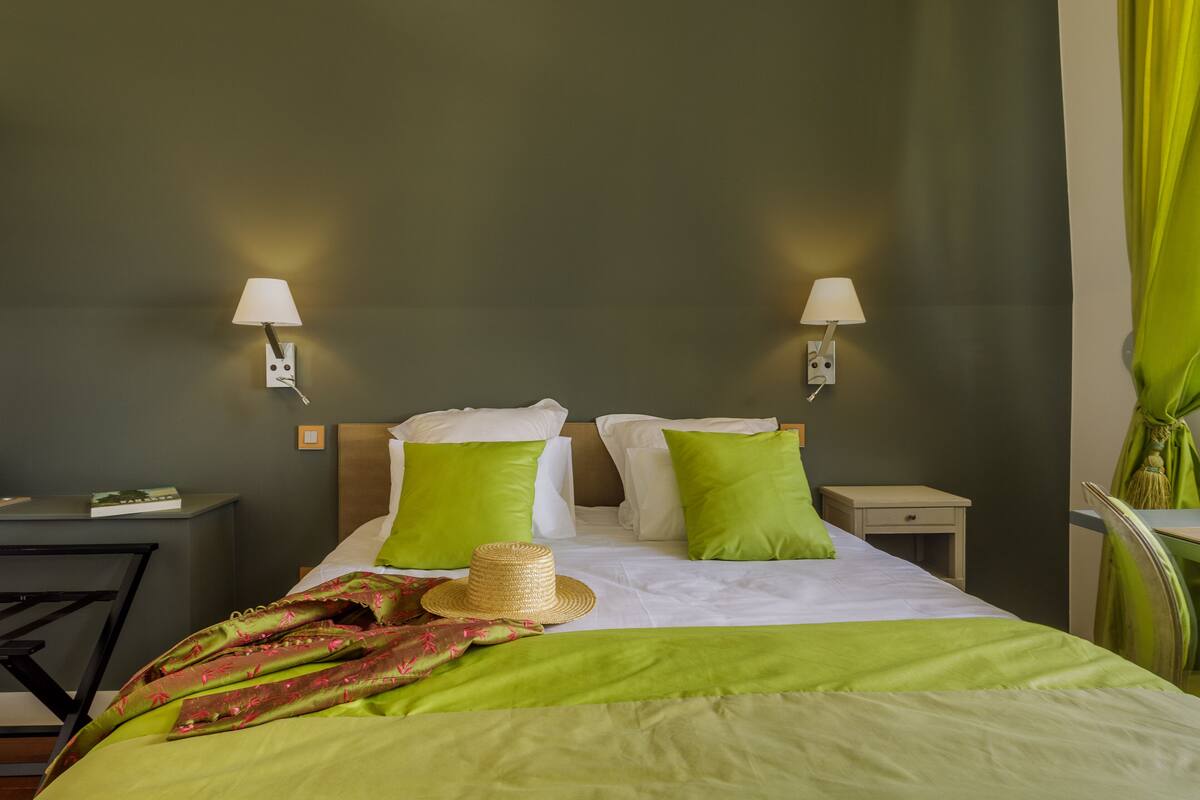 Quarto de hotel com uma cama, duas luminárias de cabeceira, uma mesa de apoio, uma escrivaninha e uma cadeira.