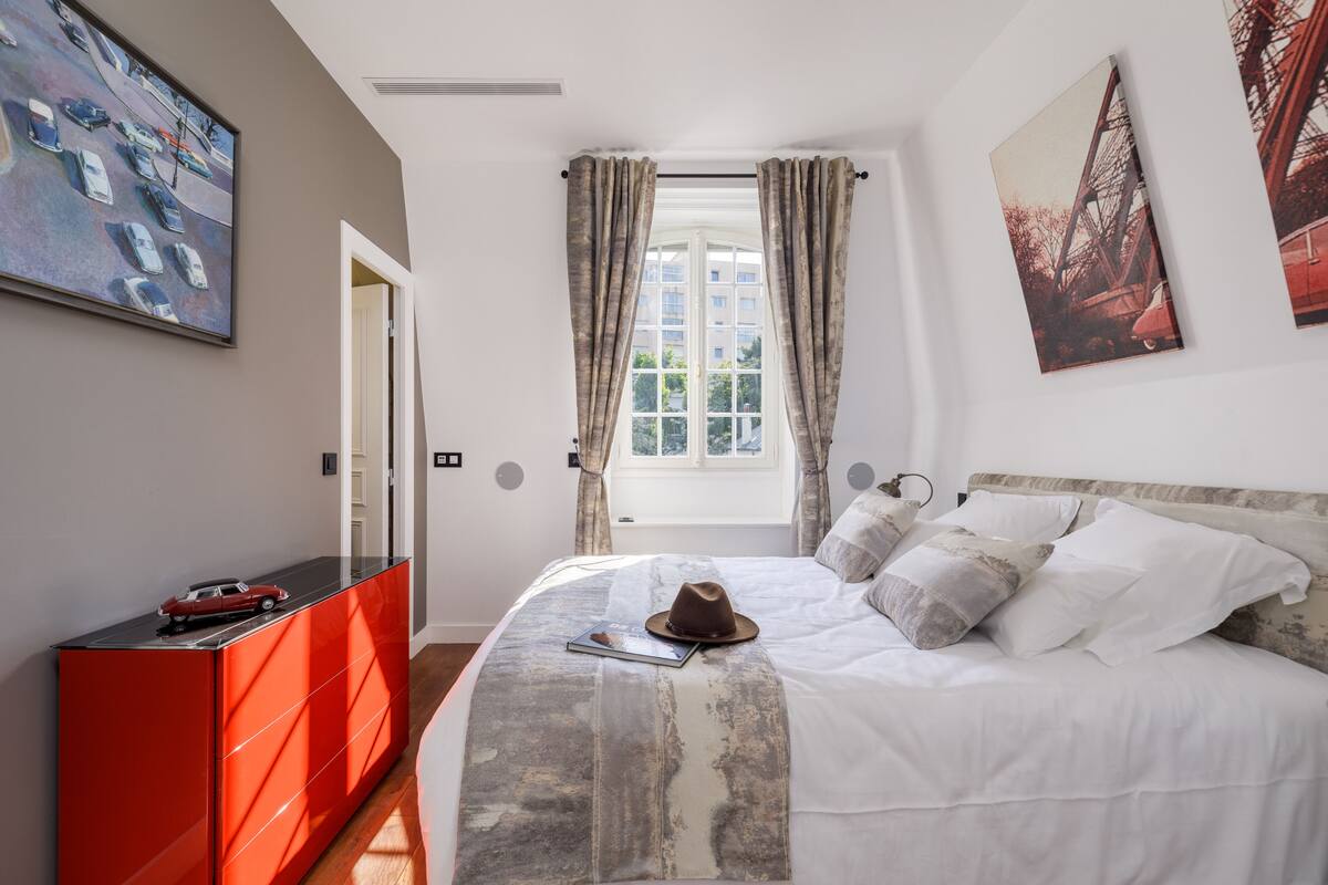 Um quarto com cama, penteadeira vermelha, janela com cortinas e obras de arte nas paredes.