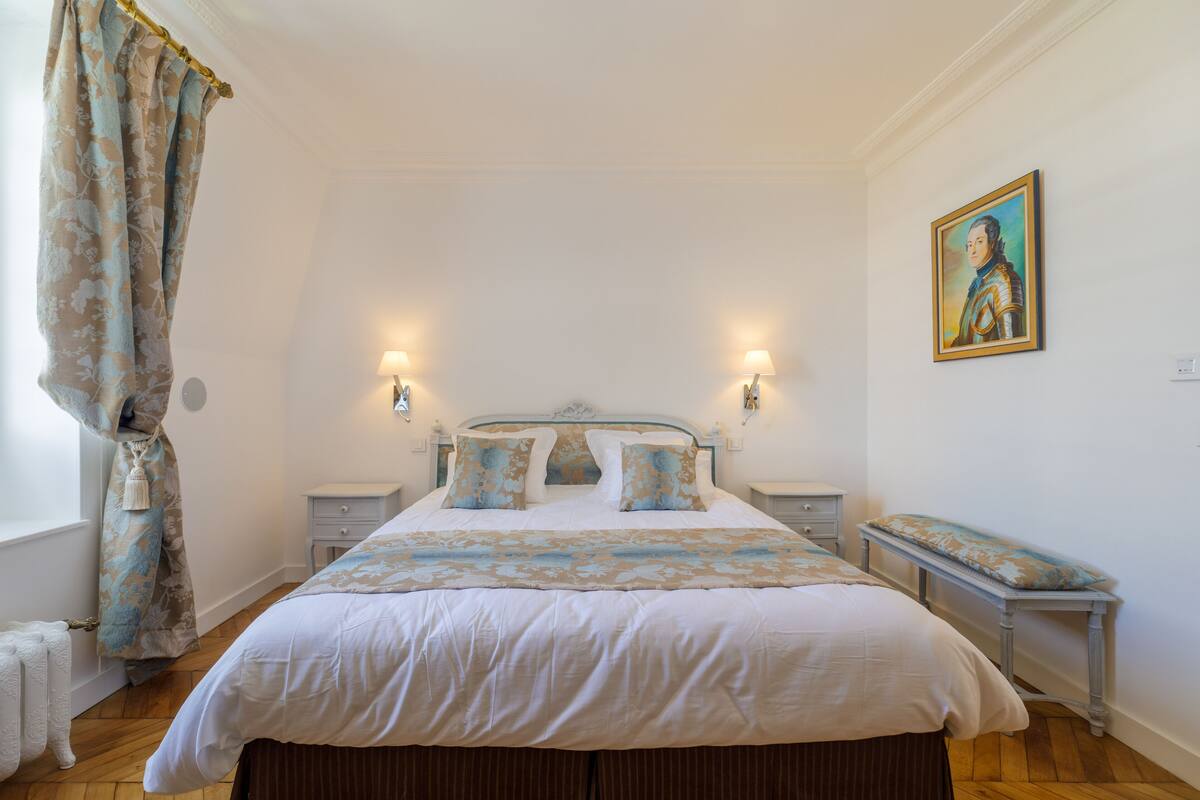 Quarto com cama grande, duas mesas de cabeceira, um banco, um quadro na parede e uma janela com cortinas.
