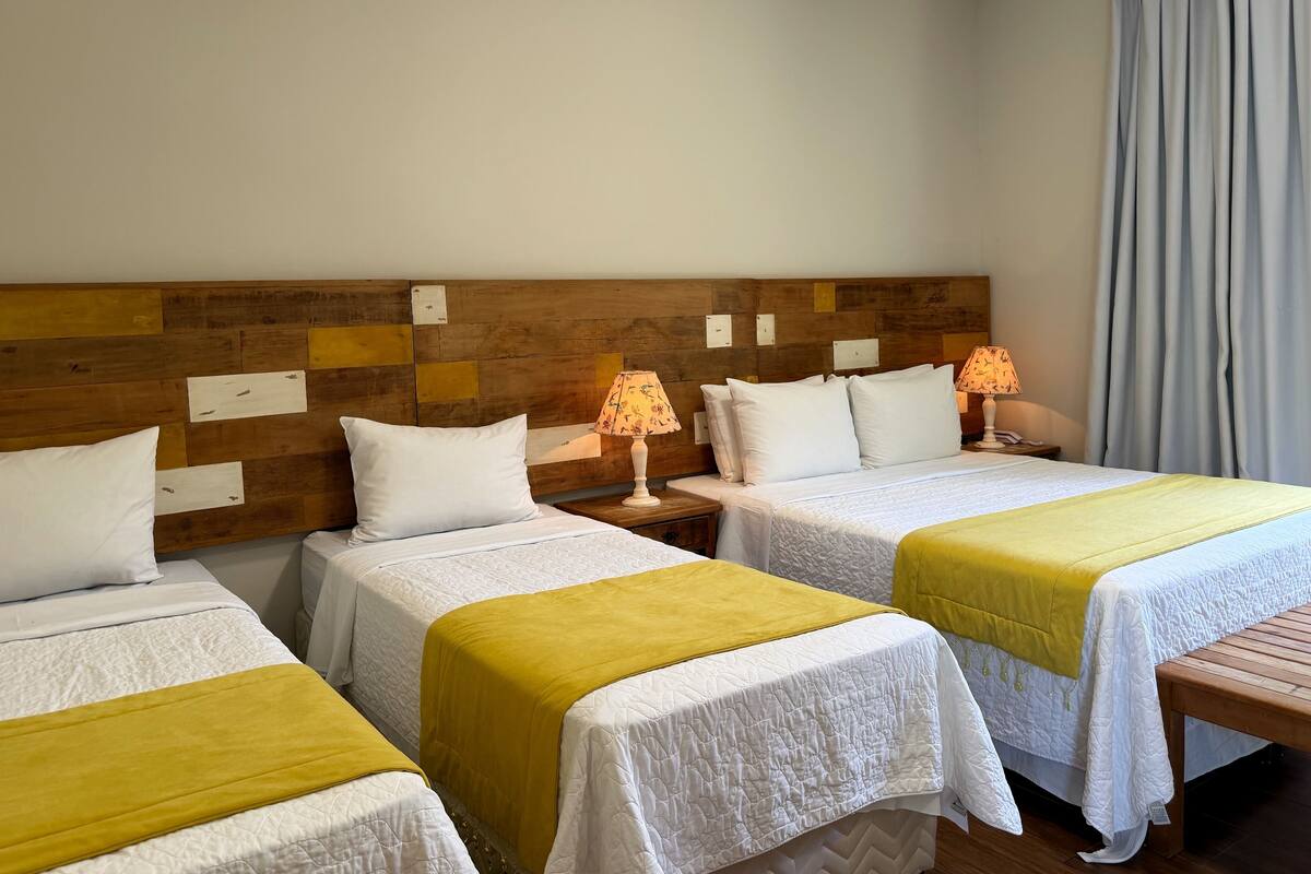 Quarto de hotel com duas camas, cabeceira de madeira, roupa de cama branca, mantas amarelas, duas luminárias de cabeceira e um banco de madeira.