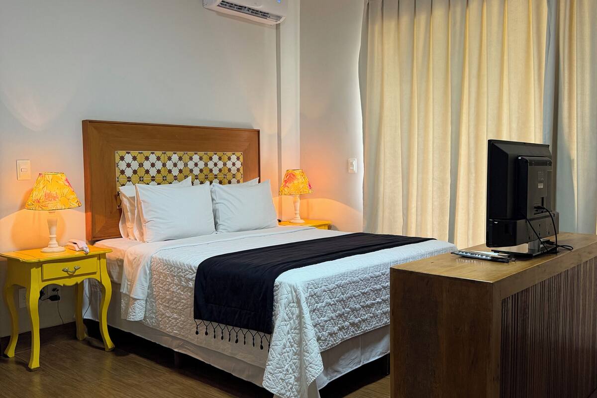 Quarto de hotel com uma cama, duas luminárias de cabeceira, uma TV sobre um suporte de madeira e uma janela com cortinas.