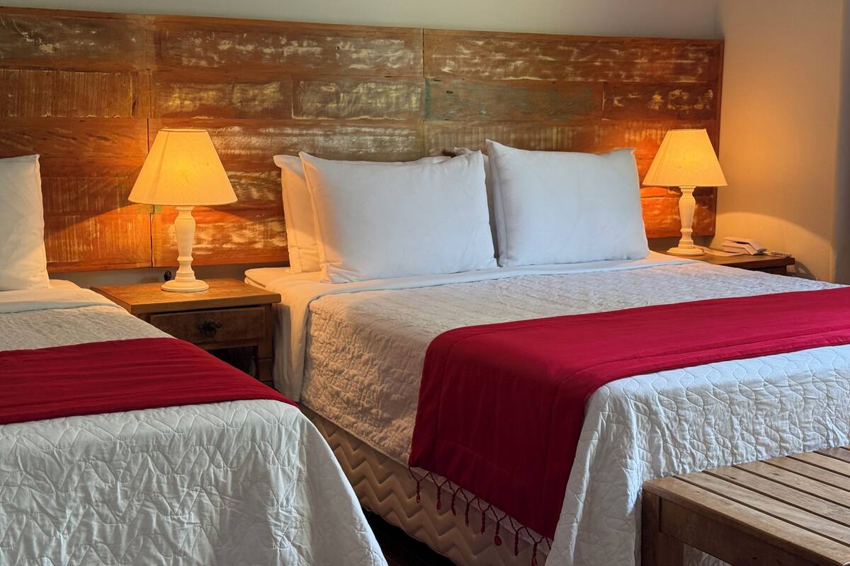 Quarto de hotel com duas camas, cada uma com roupa de cama branca e mantas vermelhas. Há duas luminárias de mesa e uma cabeceira de madeira.