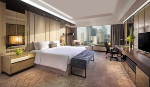 Quarto de hotel moderno com uma cama grande, uma escrivaninha, uma cadeira e uma televisão. O quarto tem vista para a cidade e uma janela ampla.