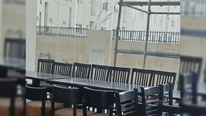 Uma área de refeições com uma mesa comprida e várias cadeiras, um corrimão e vista para edifícios ao fundo.