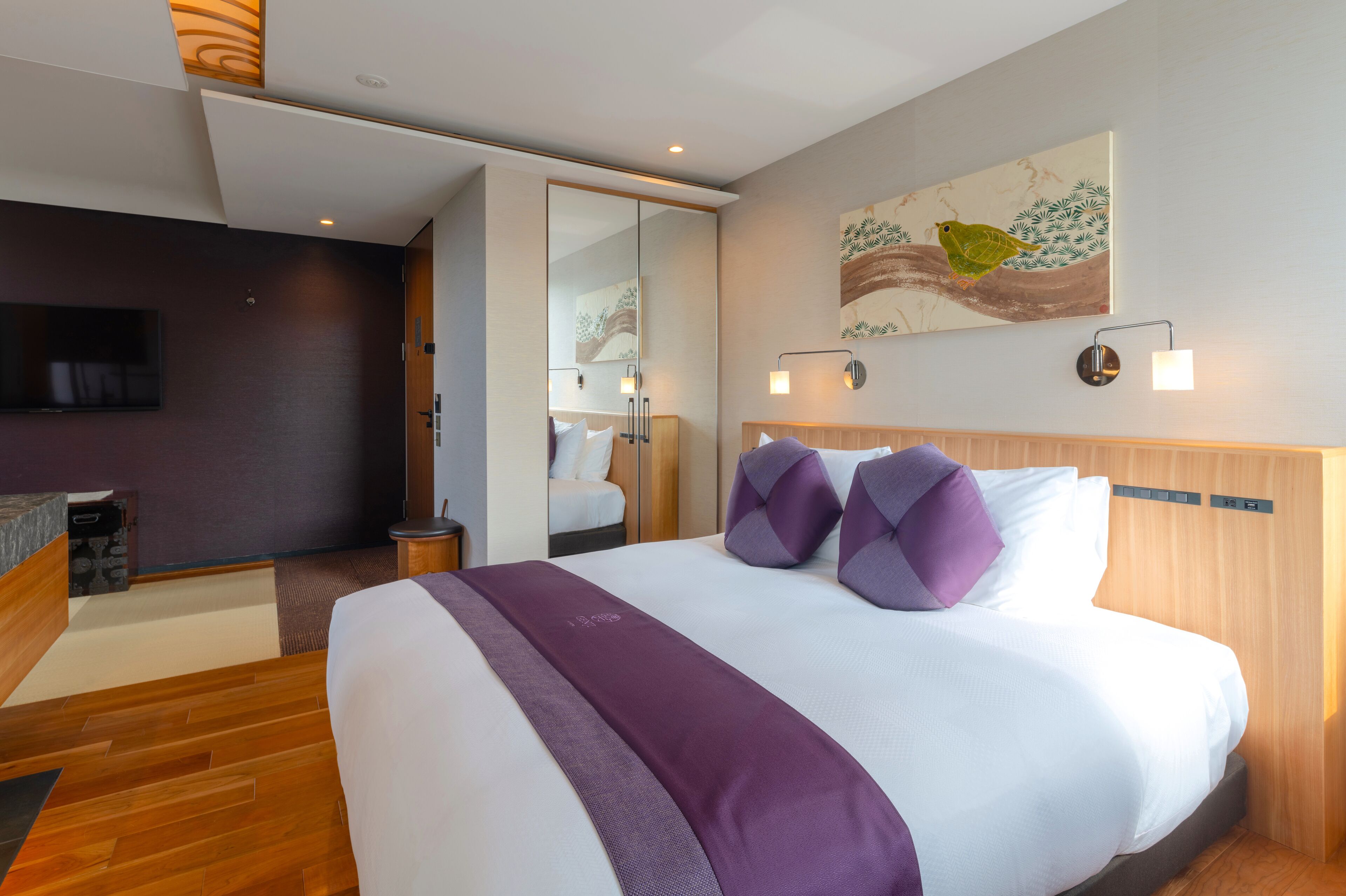 Quarto de hotel moderno com uma cama grande, televisão de tela plana, cabeceira de madeira e um quadro de um pássaro na parede.