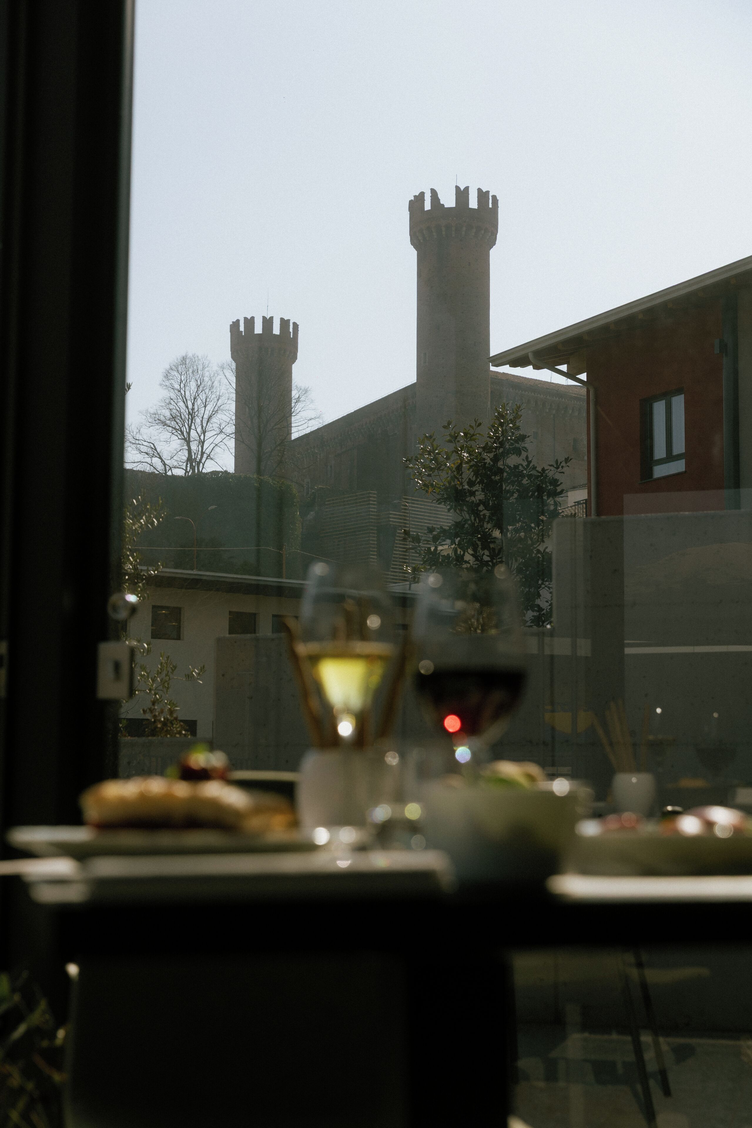 Uma mesa com um copo de vinho e um prato de comida, uma janela com vista para um edifício semelhante a um castelo.