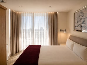 Quarto de hotel com uma cama grande, mesa de cabeceira, luminária e janela com cortinas.