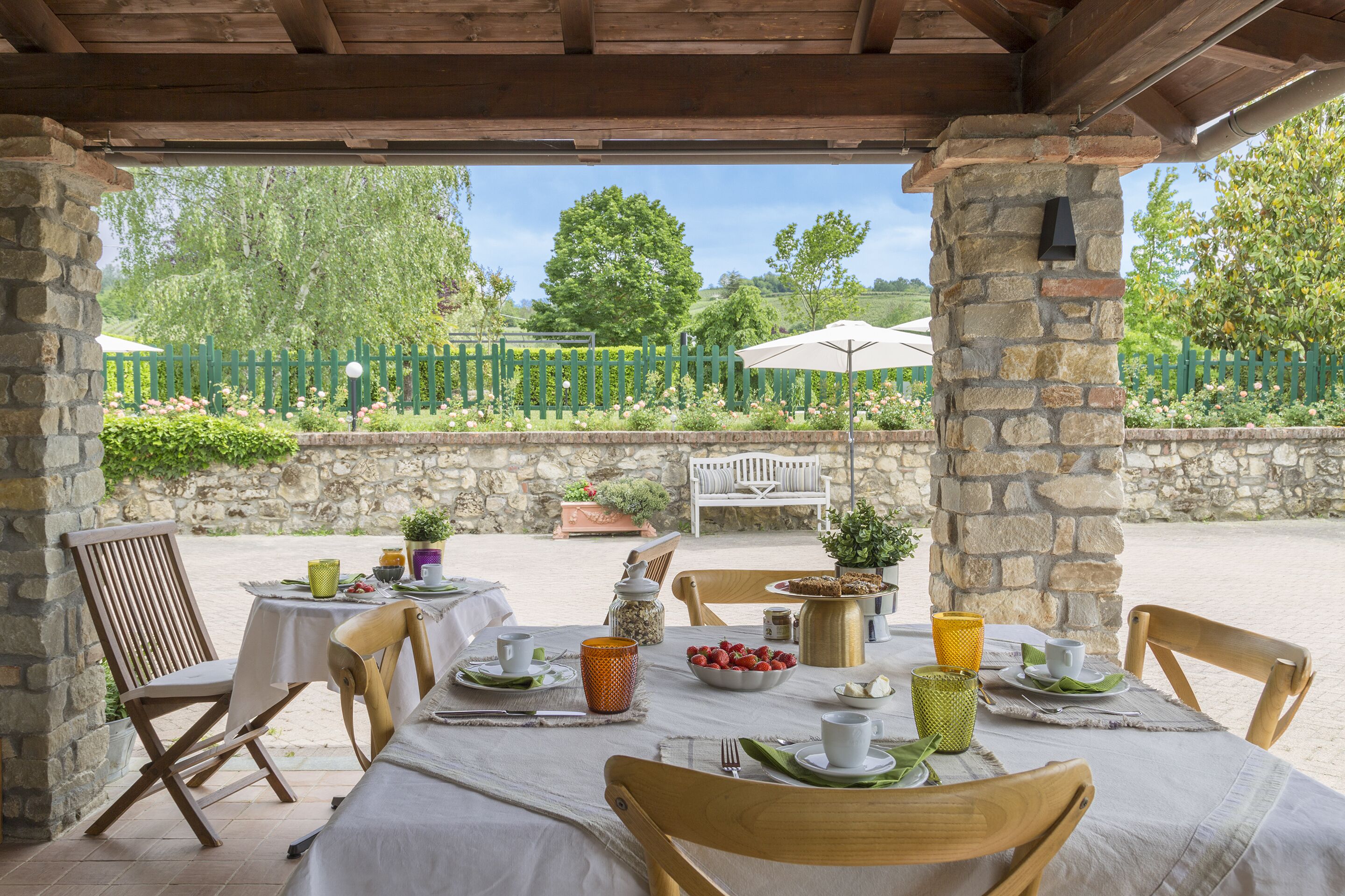 Um pátio externo com pilares de pedra, uma mesa posta para refeições e vista de um jardim com cerca verde e árvores.