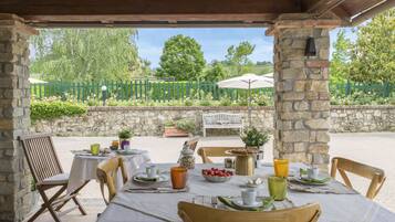 Um pátio externo com pilares de pedra, uma mesa posta para refeições e vista de um jardim com cerca verde e árvores.