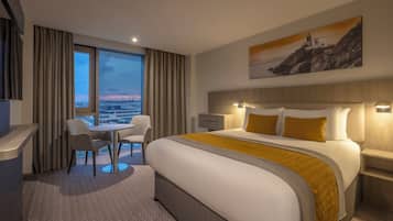 Quarto de hotel com uma cama grande, uma escrivaninha, uma cadeira e vista para o horizonte urbano através de amplas janelas.