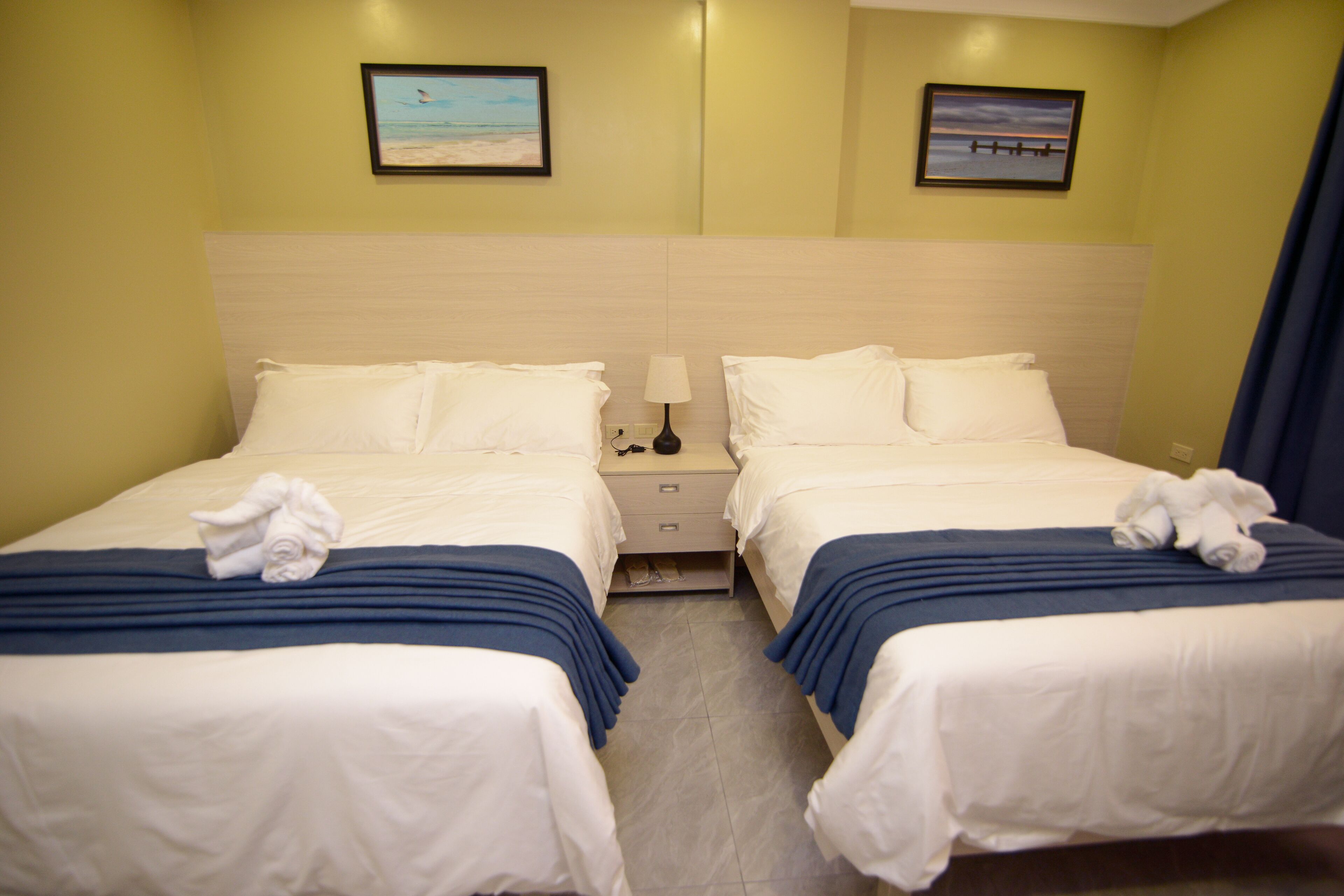Quarto de hotel com duas camas, cada uma com roupa de cama branca e detalhes azuis. Há duas imagens emolduradas nas paredes, uma retratando uma cena de praia e a outra, um pôr do sol.
