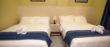 Quarto de hotel com duas camas, cada uma com roupa de cama branca e detalhes azuis. Há duas imagens emolduradas nas paredes, uma retratando uma cena de praia e a outra, um pôr do sol.