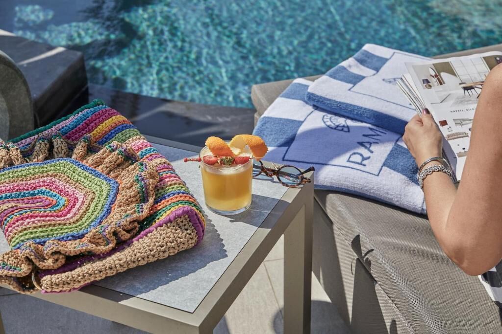Cenário à beira da piscina com um objeto colorido em crochê, uma cerveja com frutas e uma toalha com logotipo.