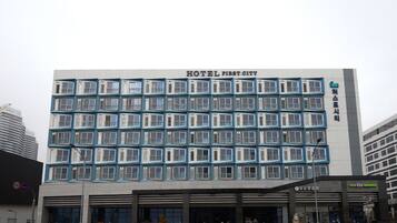 Um hotel de vários andares com fachada moderna, destacando uma grade de janelas e uma sinalização chamativa com a inscrição "HOTEL FIRST CITY".