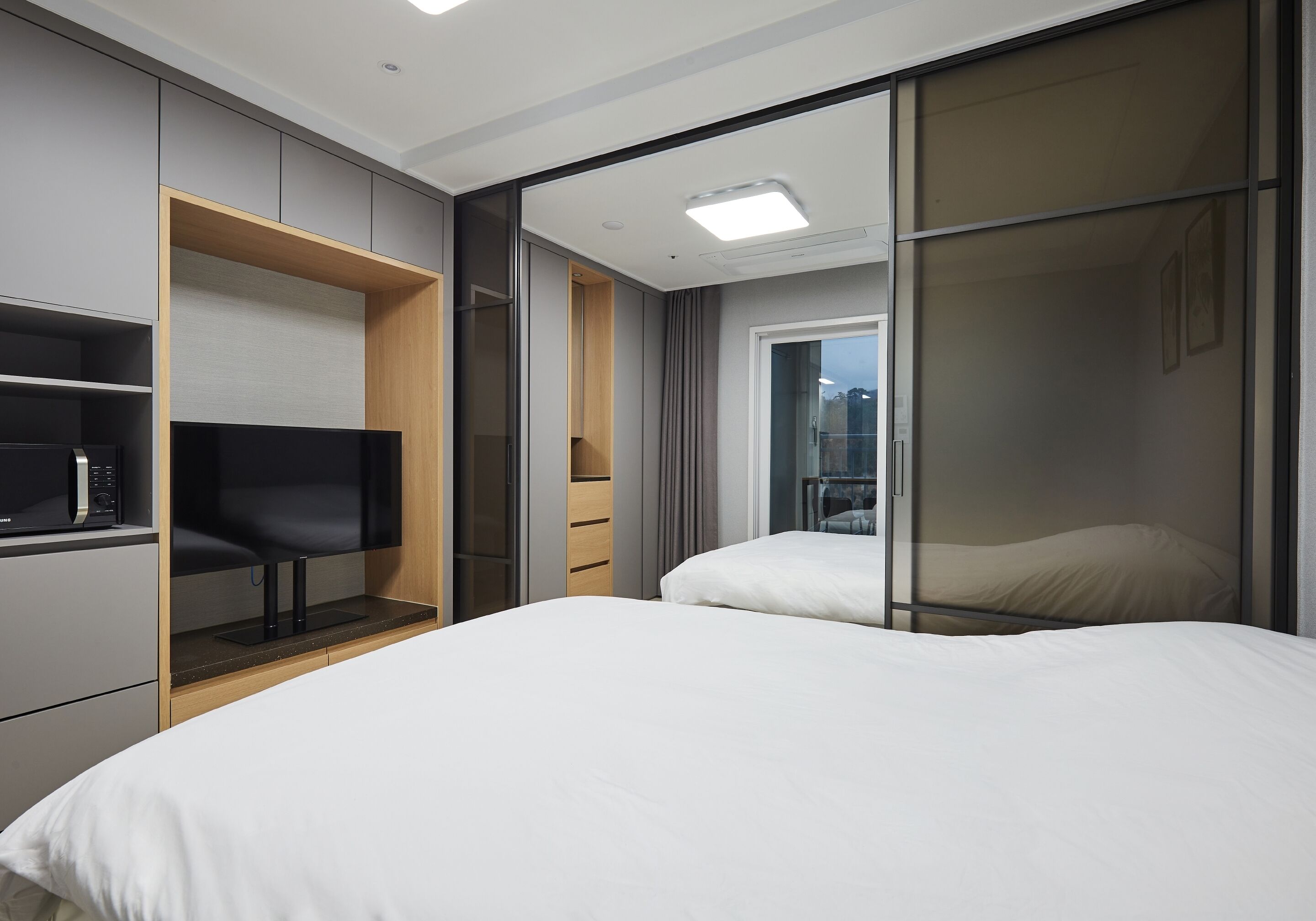 Quarto de hotel moderno com uma cama grande, televisão de tela plana, prateleiras embutidas e uma porta de vidro deslizante para uma varanda.