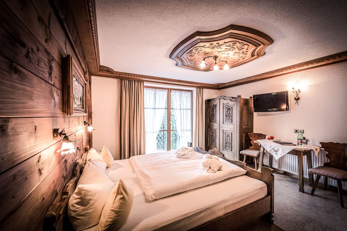 Quarto de hotel com uma cama grande, painéis de madeira, um candelabro, uma televisão e uma mesa com toalha.