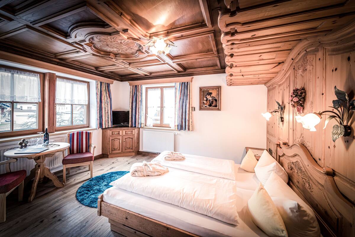 Um quarto acolhedor com teto de madeira, uma cama com lençóis brancos, uma mesa pequena com uma cadeira e uma janela com cortinas.