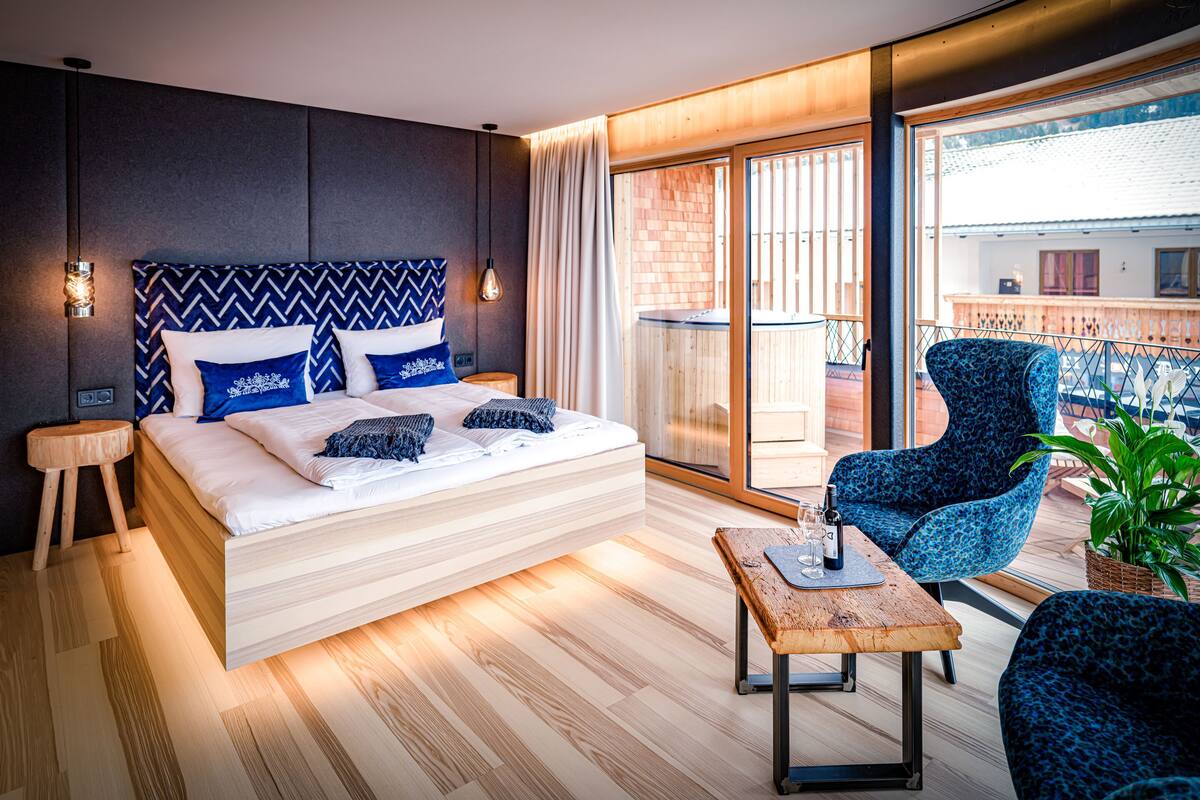 Quarto de hotel moderno com uma cama grande, uma mesinha de madeira, uma poltrona azul e uma varanda com vista para a cidade.