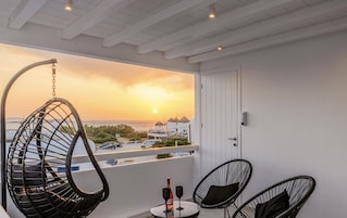 Uma varanda com uma cadeira suspensa, duas cadeiras, uma mesa com garrafa e copos, e vista para o pôr do sol sobre o mar.