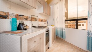 Cozinha com fogão, forno e diversos eletrodomésticos. Há uma janela com cortinas e vista para prédios.