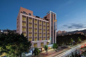 Um hotel de vários andares com design moderno, destacando um elemento vertical proeminente no lado direito, e rodeado por árvores e uma rua movimentada.