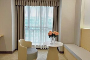 Quarto de hotel com uma janela grande, uma mesa redonda, um sofá e um vaso com flores.
