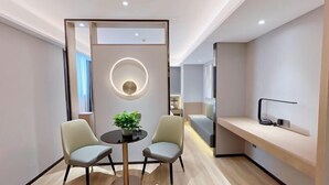 Quarto de hotel moderno com uma mesa redonda, duas cadeiras, um vaso de plantas, uma escrivaninha com abajur e uma prateleira na parede.