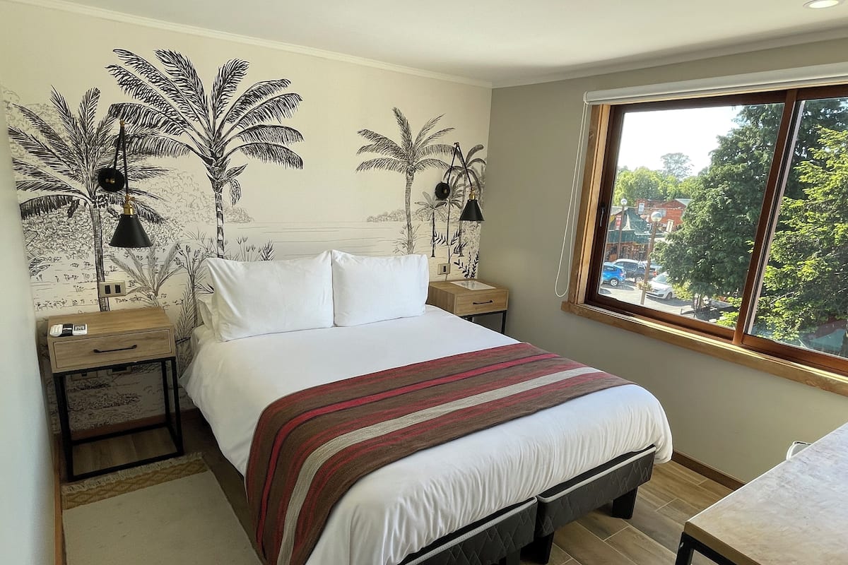 Quarto bem arrumado com uma cama grande, duas mesas de cabeceira, um criado-mudo, uma janela com vista para a rua e quadros de palmeiras na parede.