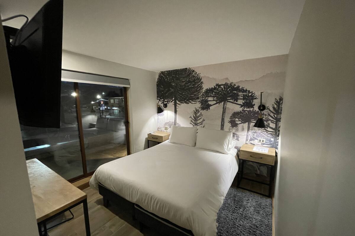 Quarto com cama, mesas de cabeceira, escrivaninha e uma janela grande.