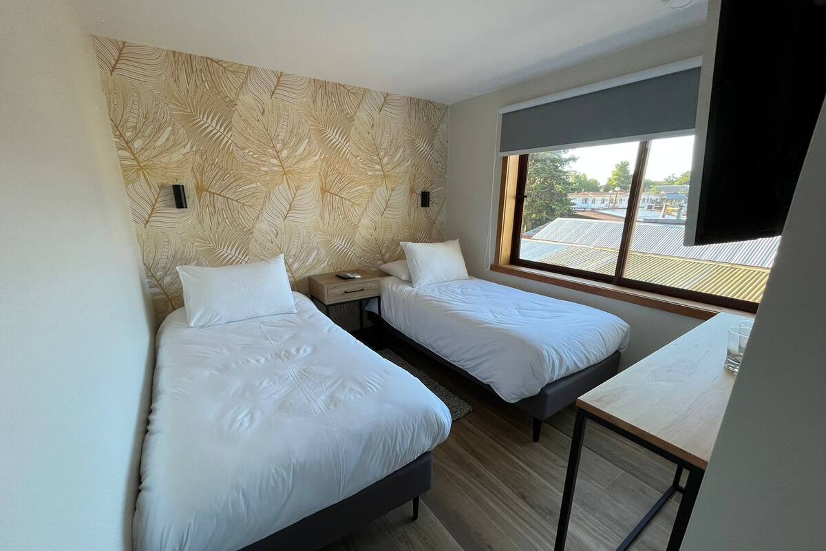 Quarto de hotel moderno com duas camas de solteiro, uma mesinha, um criado-mudo e uma janela grande com vista para o exterior.