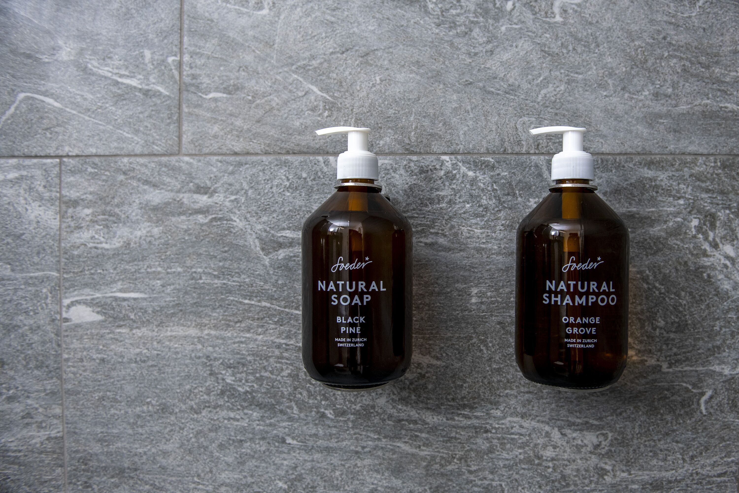Duas garrafas de sabonete e shampoo natural encostadas a uma parede azulejada.