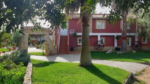 Um edifício vermelho com entrada de pedra, um caminho pavimentado e um jardim bem cuidado com diversas plantas.