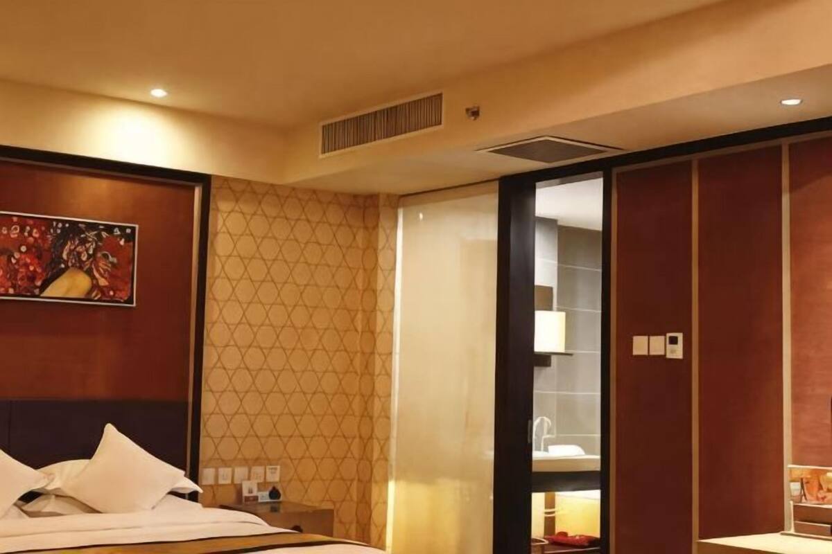 Quarto de hotel com cama, mesa de cabeceira, obra de arte na parede, papel de parede estampado e porta deslizante que dá para um banheiro.