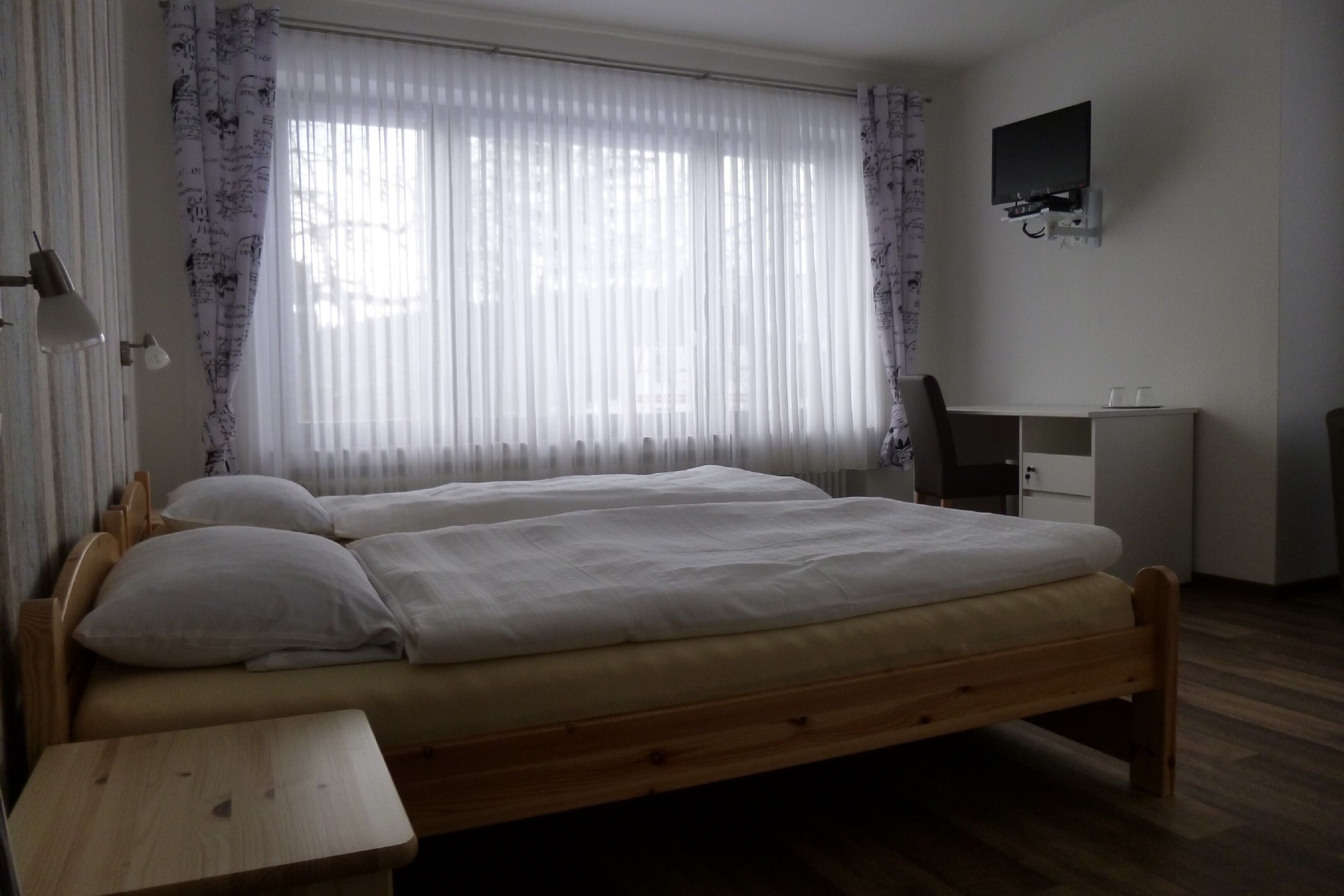 Quarto com duas camas de solteiro, uma escrivaninha, uma cadeira e televisão fixada na parede.
