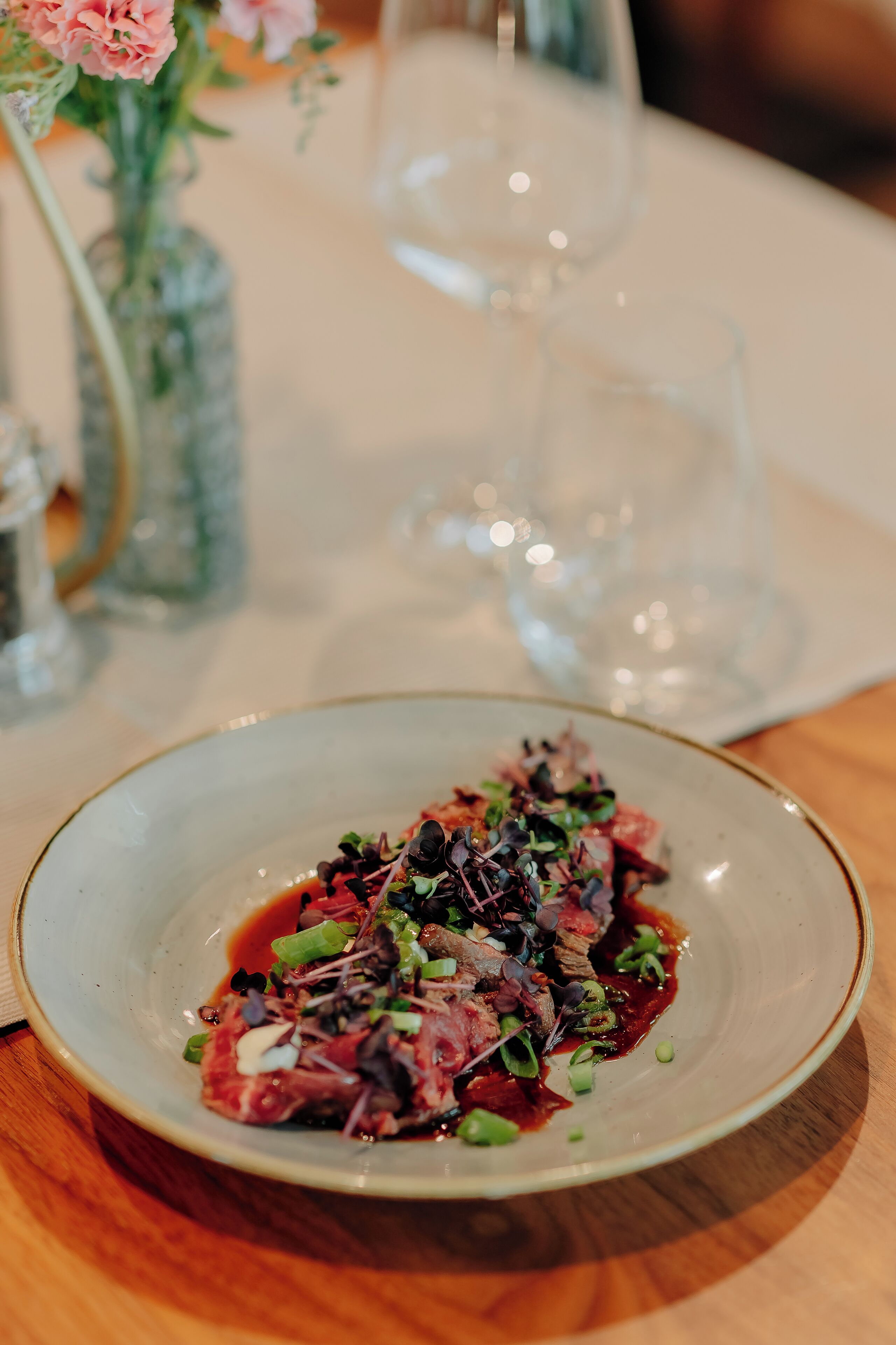 Prato de comida com diversos ingredientes, incluindo vegetais e possivelmente carne, sobre uma mesa com um vaso de flores e copos de água ao fundo.
