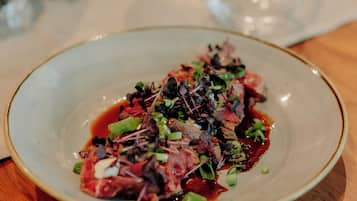 Prato de comida com diversos ingredientes, incluindo vegetais e possivelmente carne, sobre uma mesa com um vaso de flores e copos de água ao fundo.