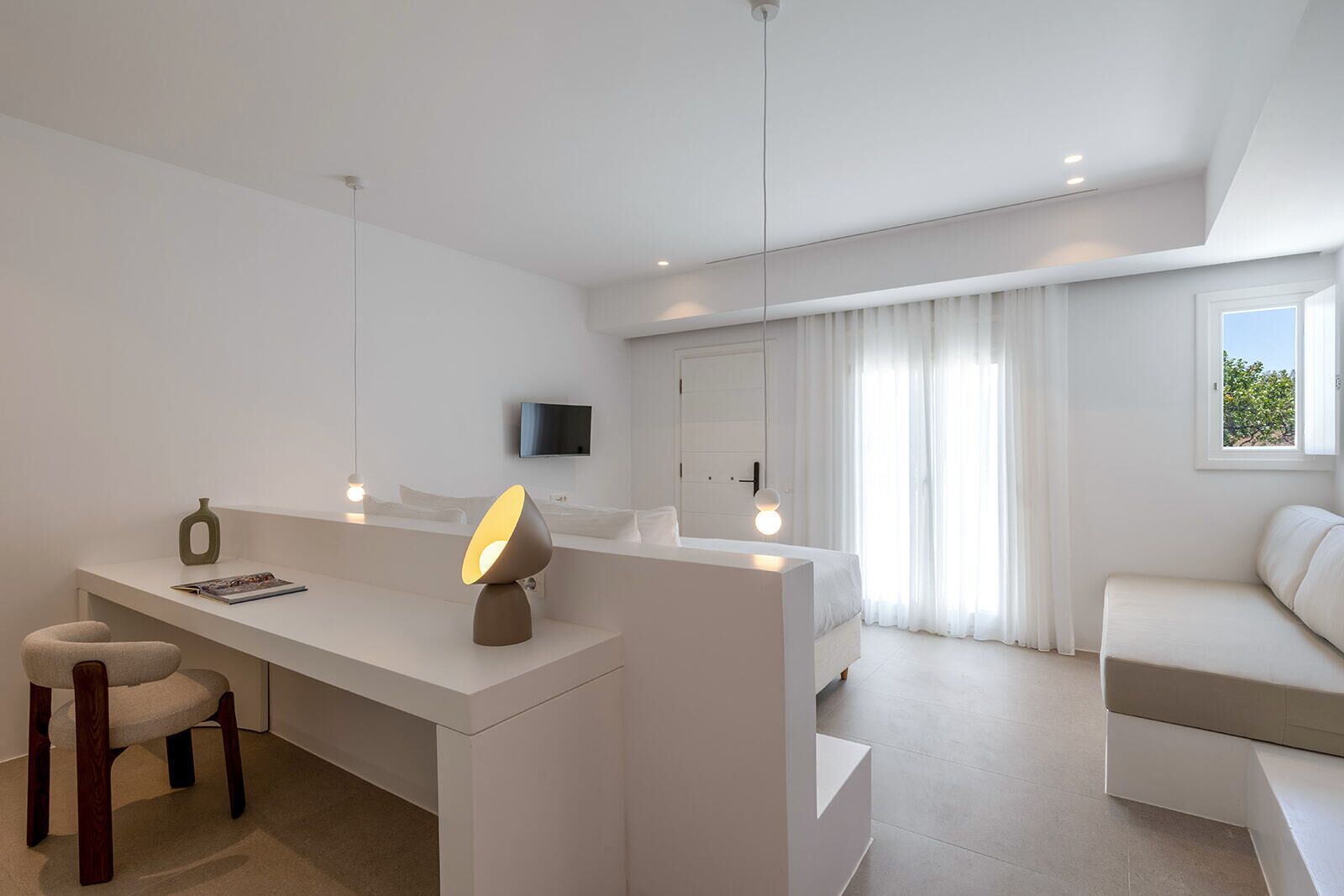 Quarto de hotel moderno com escrivaninha branca, sofá, cama e televisão.