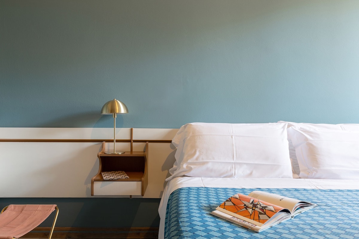 Quarto de hotel com cama, mesa de cabeceira, abajur e revista na cama.