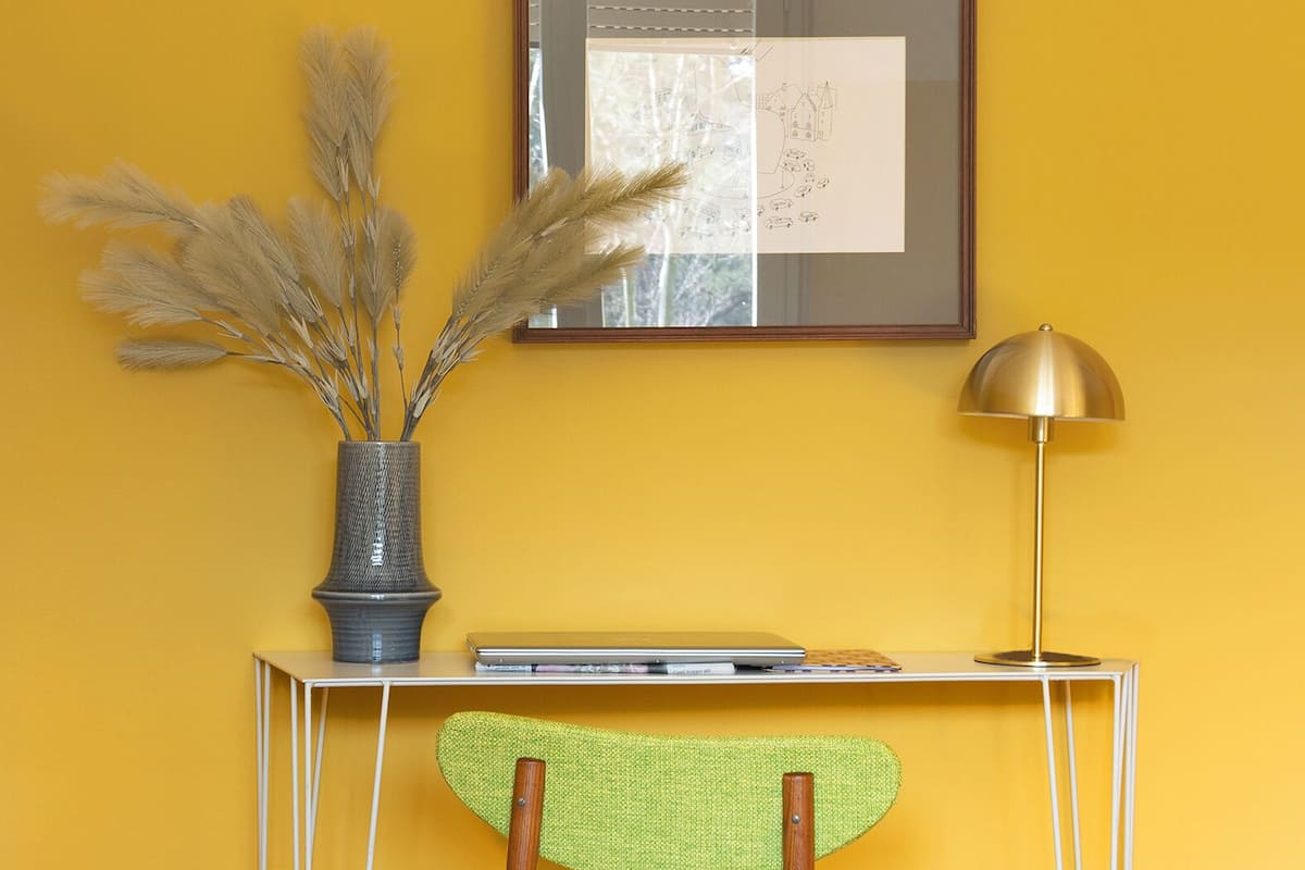 Quarto com parede amarela, quadro emoldurado, escrivaninha com laptop, cadeira verde e um vaso com plumas de capim-do-campo.