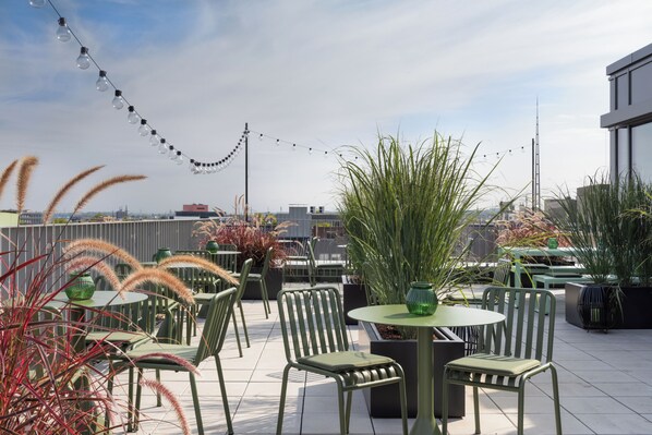 Terraço ao ar livre com cadeiras e mesas verdes, rodeado por plantas em vasos e luzes decorativas.